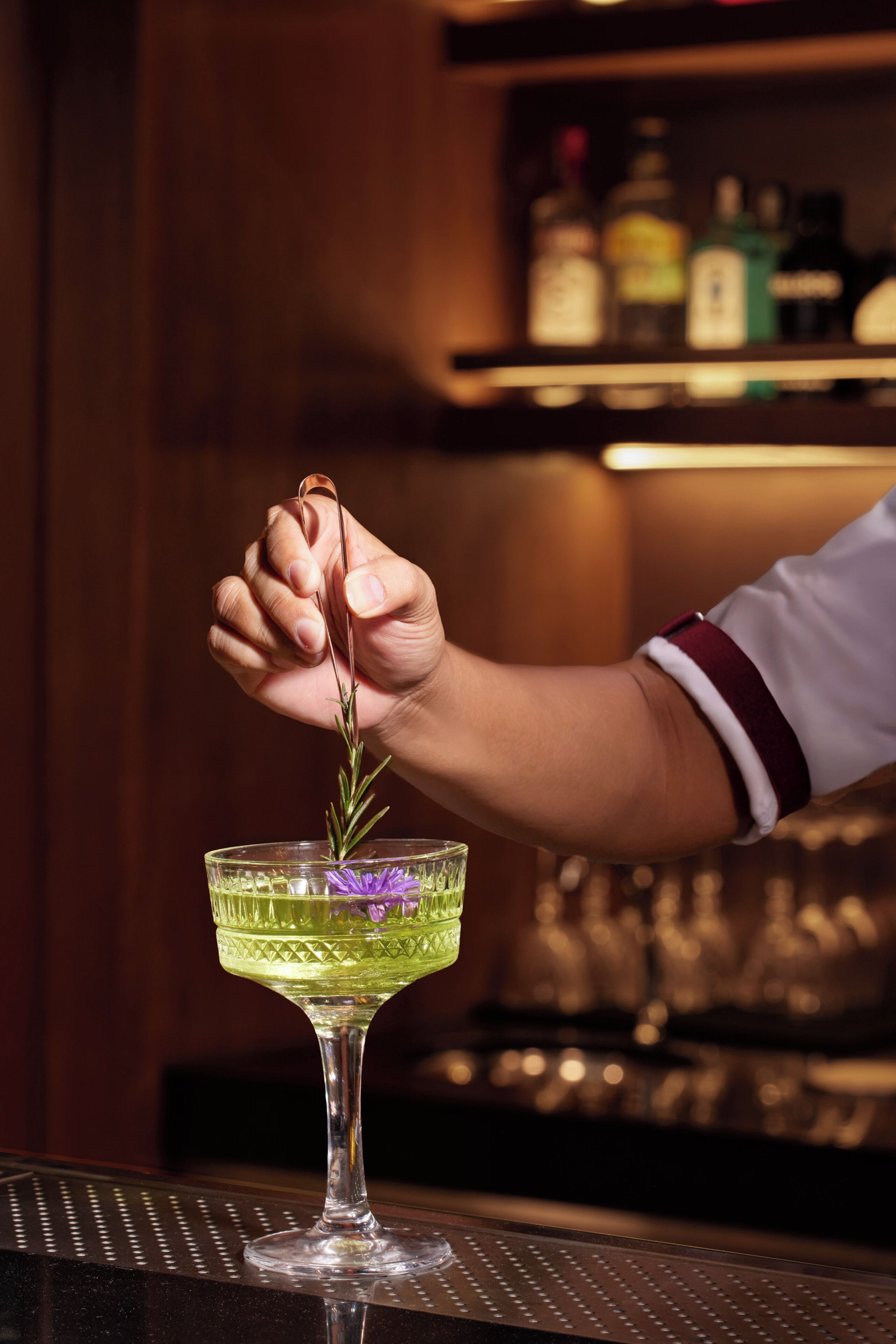 Mão de bartender segurando um ramo de alecrim sobre um copo de coquetel.