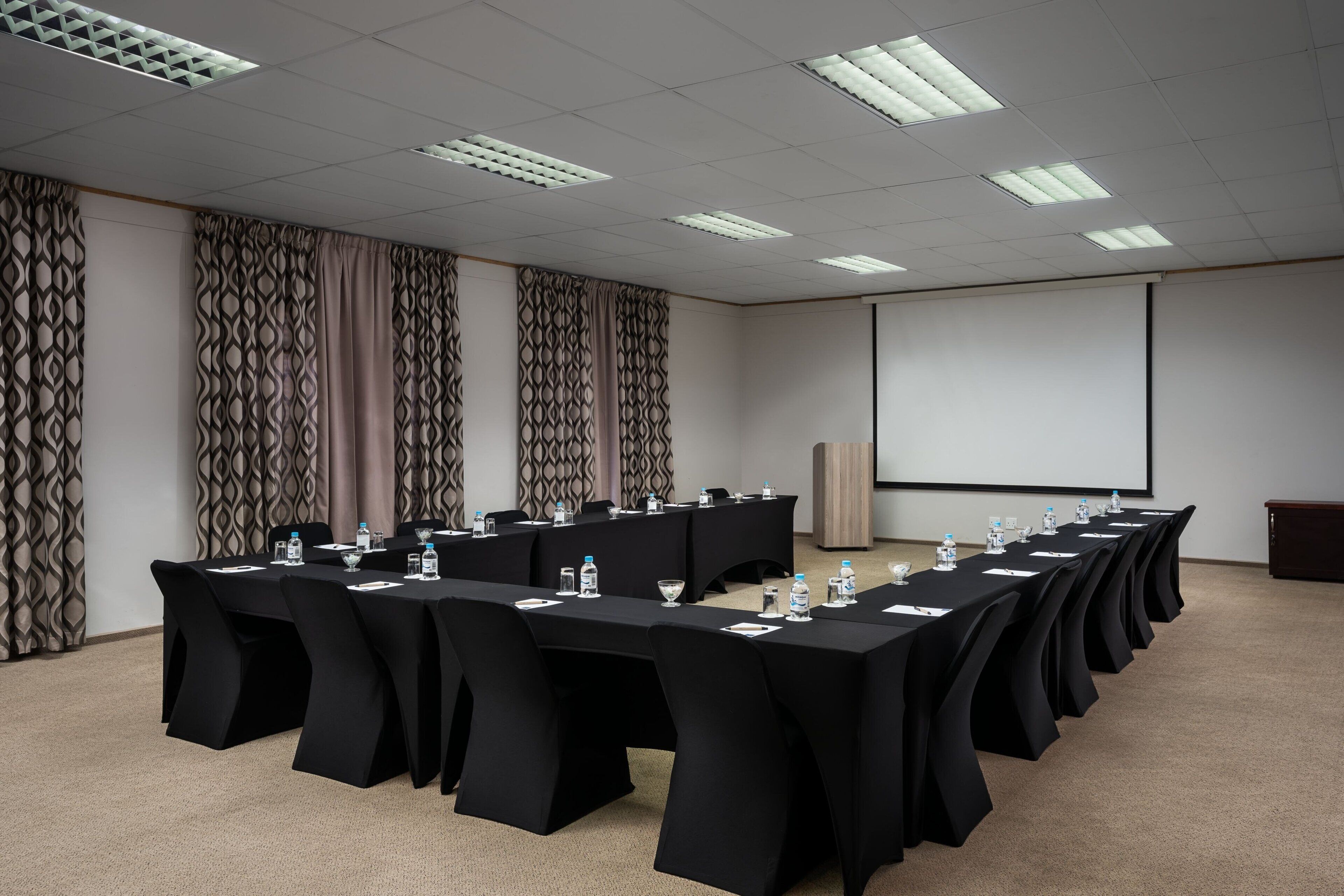 Uma sala de conferências com mesas compridas, cadeiras e uma tela de projetor.