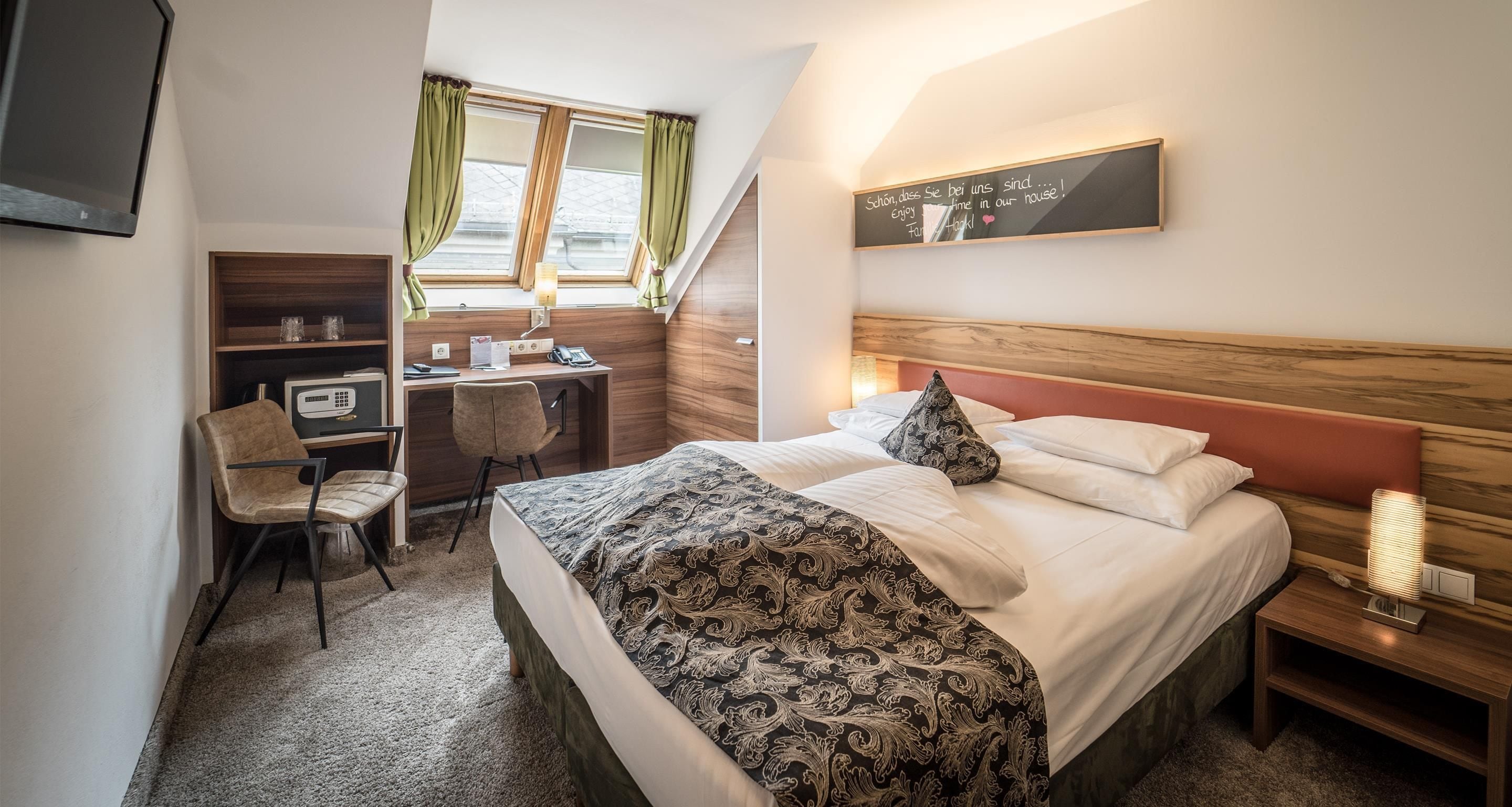 Quarto de hotel com cama, escrivaninha, cadeira e televisão fixada na parede.