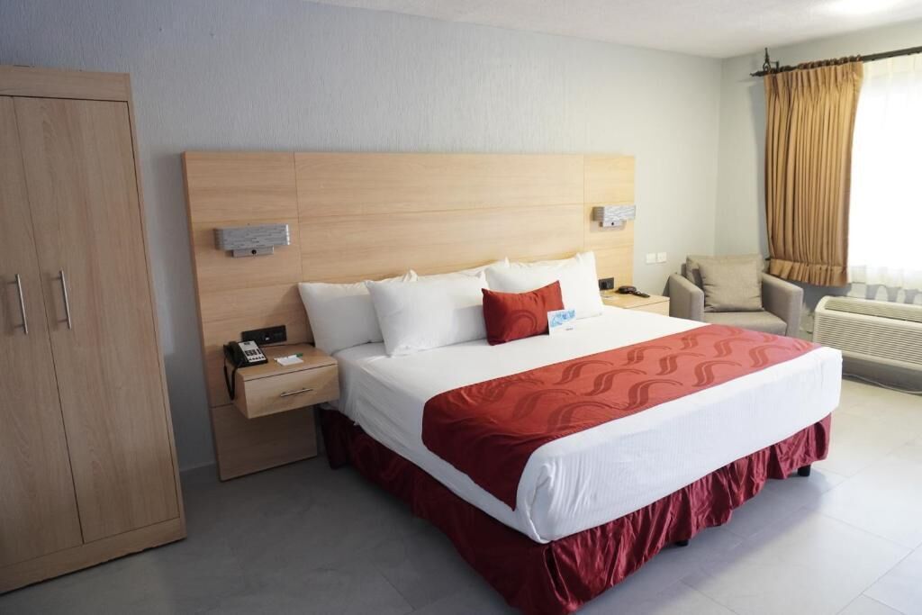 Quarto de hotel com uma cama grande, um guarda-roupa de madeira, uma mesa de cabeceira com telefone e uma cadeira.