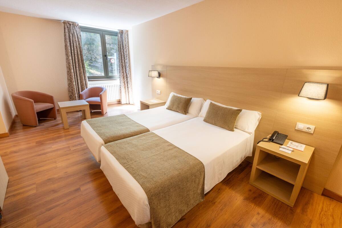 Quarto de hotel com duas camas, uma cadeira, uma mesinha, uma mesa de cabeceira e uma janela com cortinas.