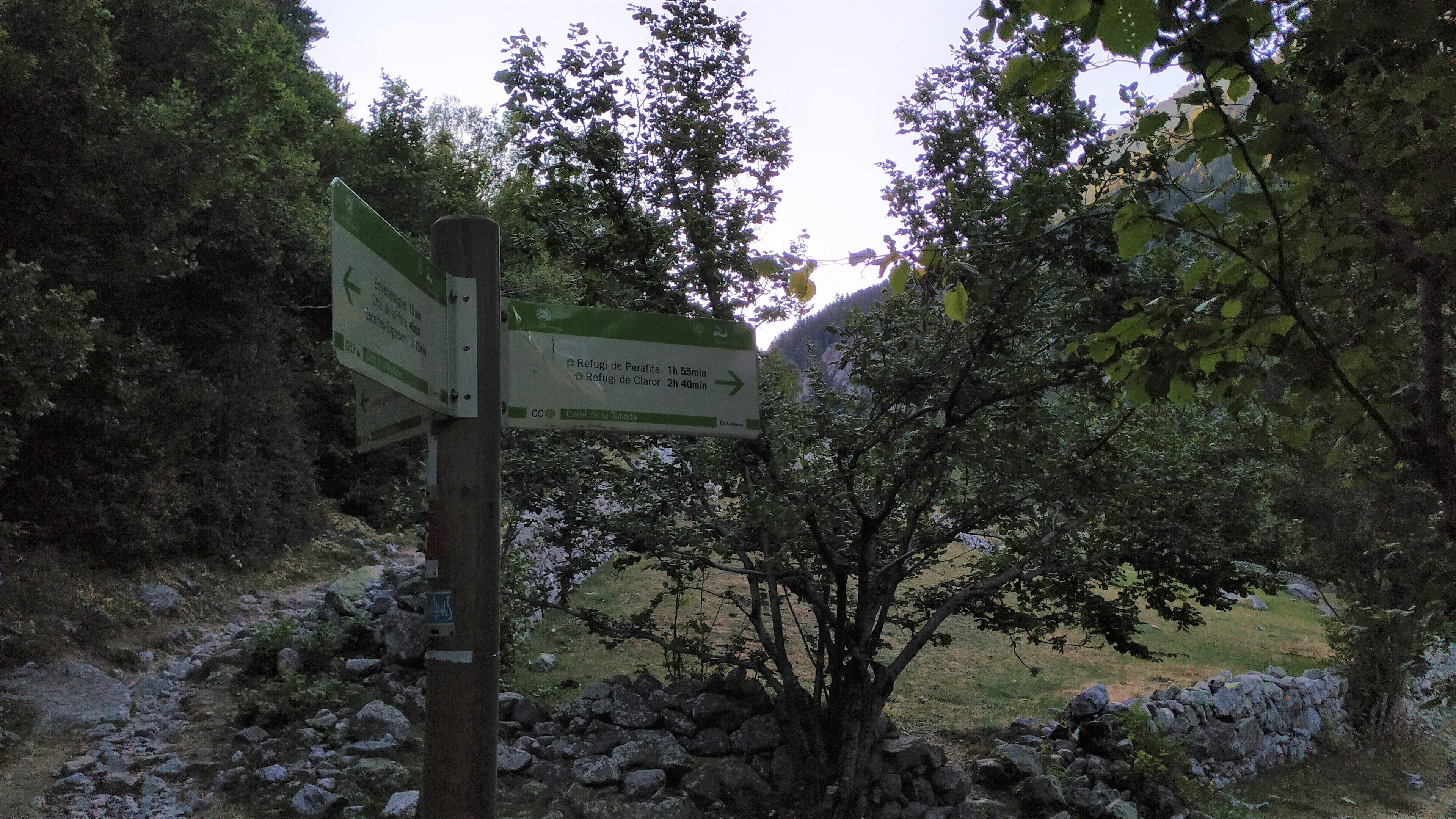 Uma placa de direção em ambiente natural, com um muro de pedra e árvores.