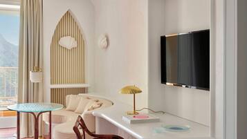 Quarto moderno com televisão de tela plana na parede, mesa de vidro redonda, duas cadeiras de vime, um sofá e uma pequena mesa de leitura com abajur.