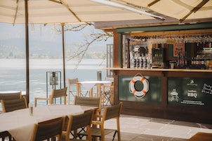 Uma área de bar ao ar livre com vista para um lago, mesas e cadeiras de madeira, e uma decoração com boia salva-vidas.