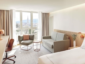 Quarto de hotel com cama, dois sofás, escrivaninha, cadeira, mesinha de centro e vista para a cidade.