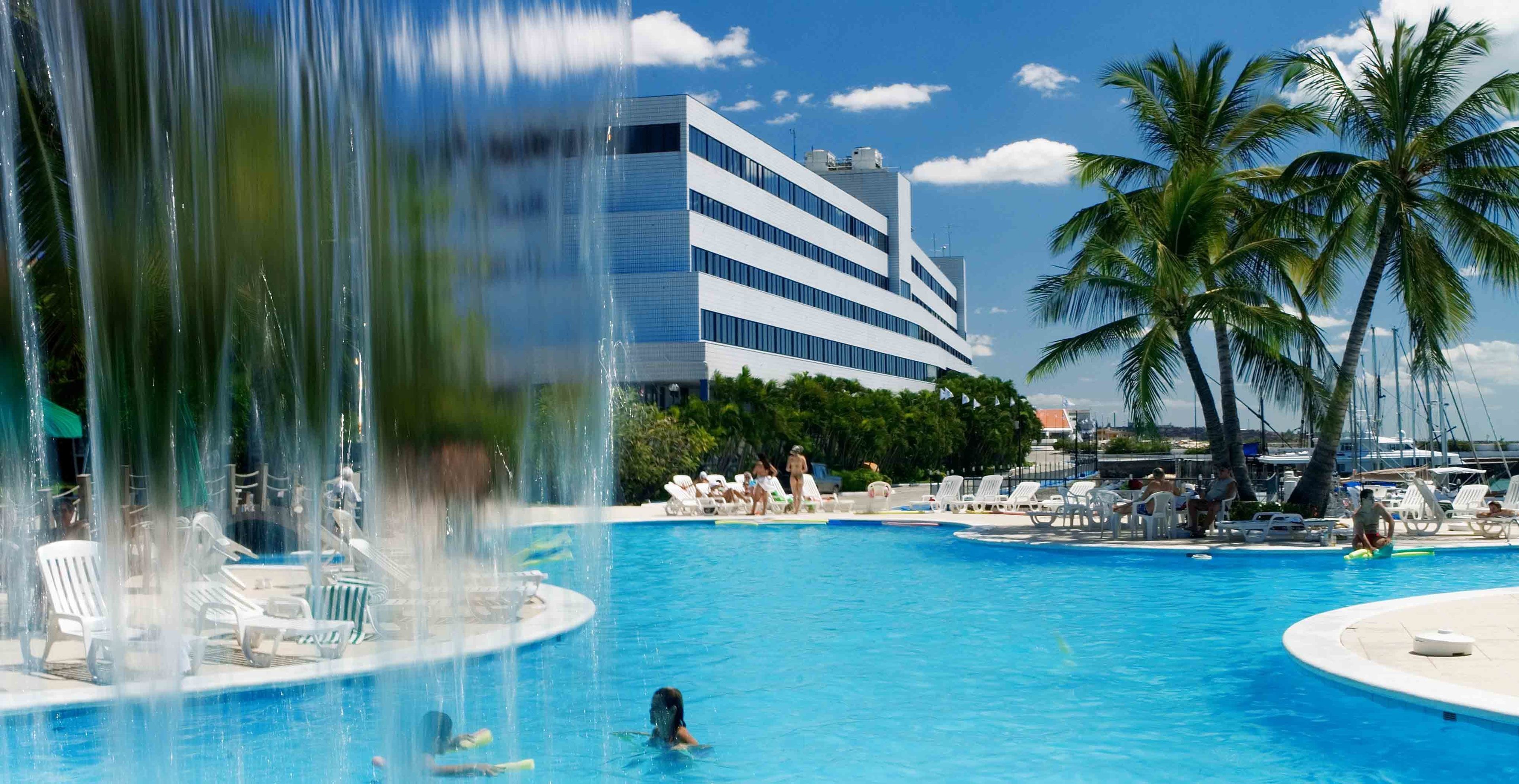 Uma piscina com uma fonte, cadeiras de descanso, palmeiras e um edifício moderno ao fundo.