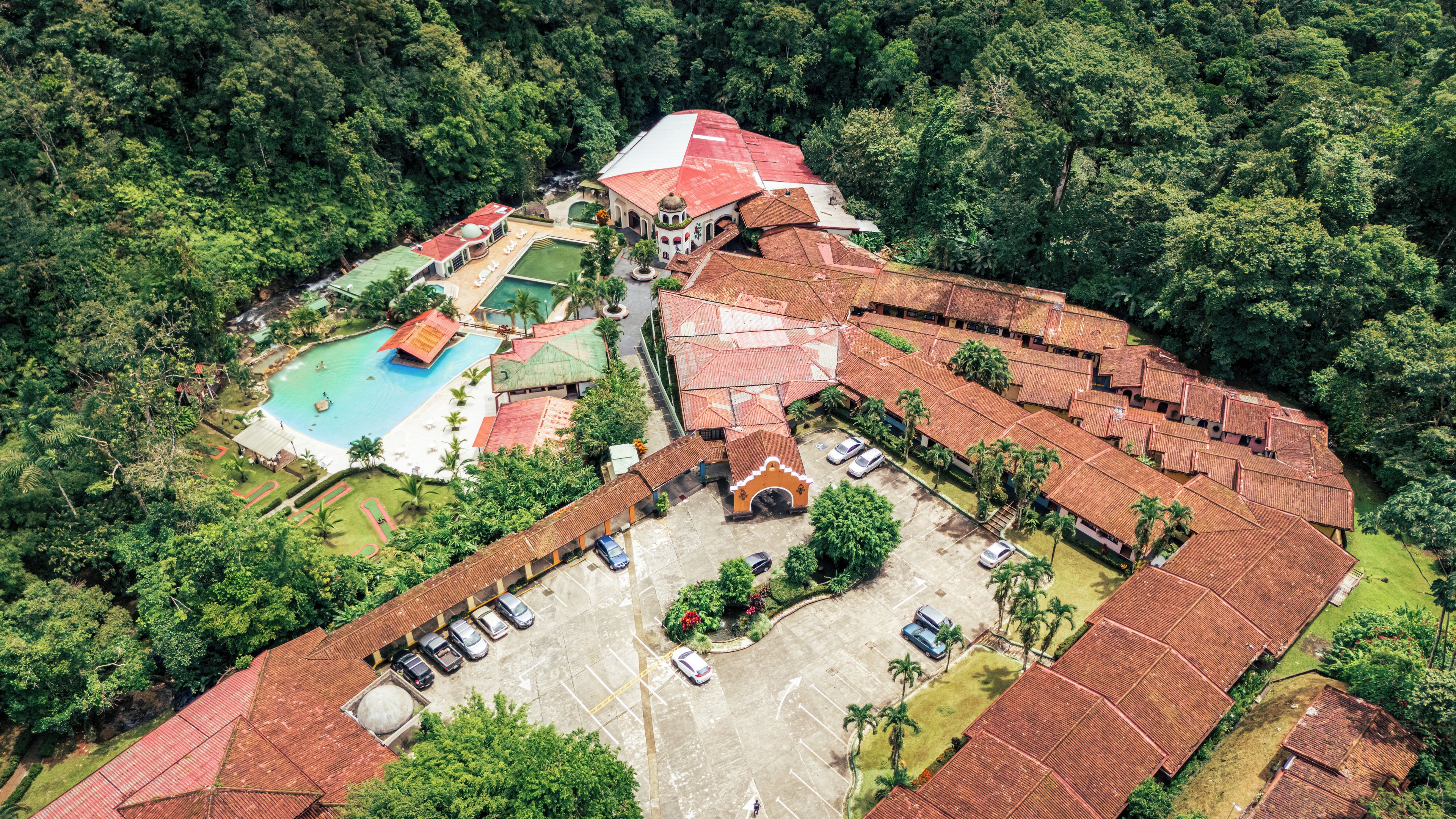 Vista aérea de um resort com vários edifícios, piscina e estacionamento, tudo cercado por vegetação exuberante.
