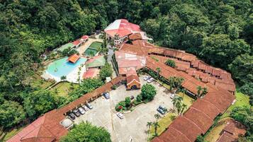 Vista aérea de um resort com vários edifícios, piscina e estacionamento, tudo cercado por vegetação exuberante.