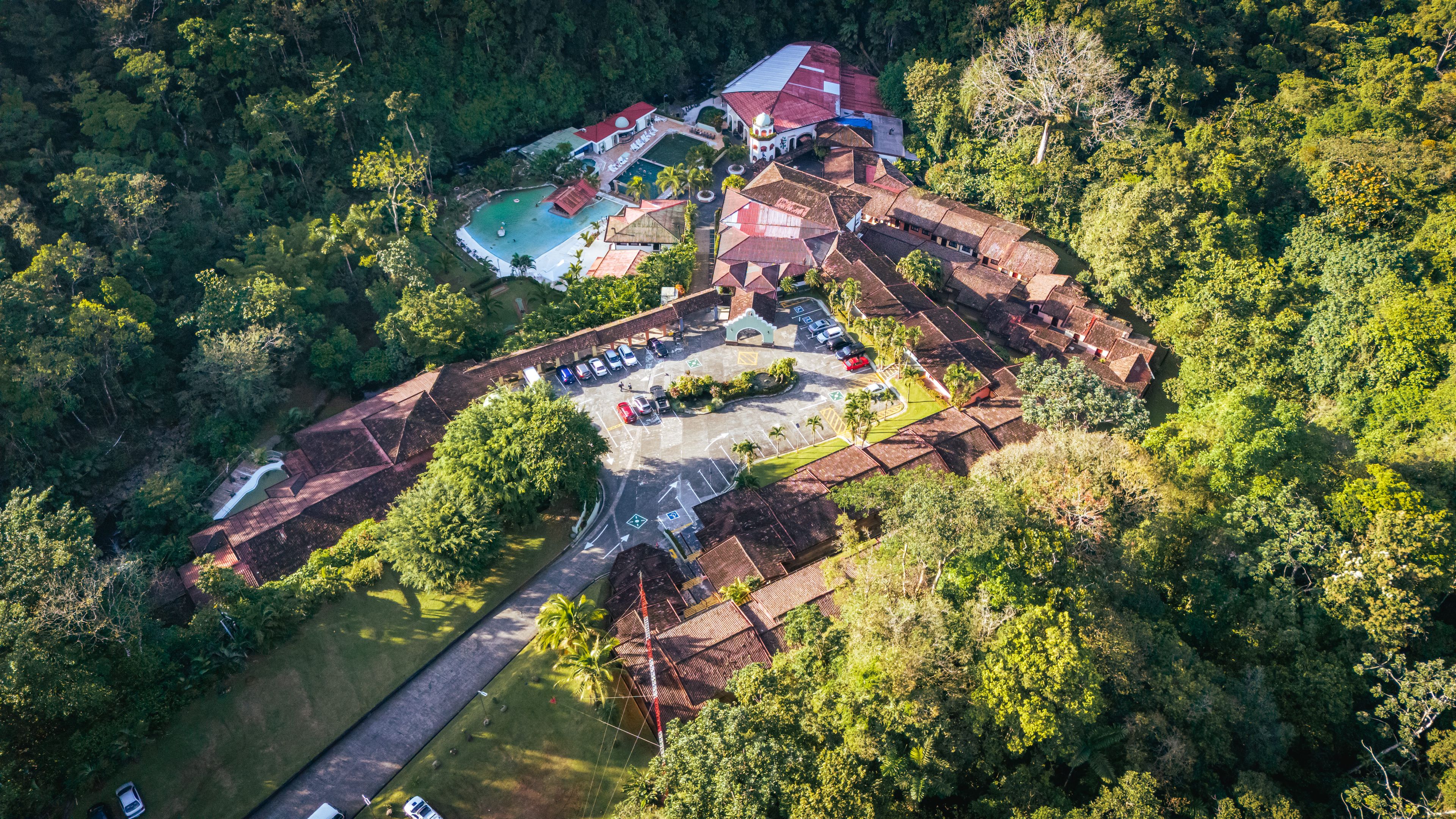 Vista aérea de um resort com vários edifícios, piscina e estacionamento, tudo cercado por uma floresta densa.