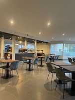 Um café moderno com várias mesas e cadeiras, um balcão com máquina de café e amplas janelas que proporcionam luz natural.