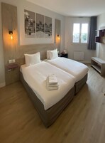 Uma cama de casal com lençóis brancos e uma toalha dobrada, um criado-mudo, uma luminária na parede e uma janela com cortinas.