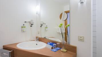 Casa de banho com uma pia branca, um espelho e bancada de madeira. Há artigos de higiene pessoal e um vaso com plantas verdes na bancada.
