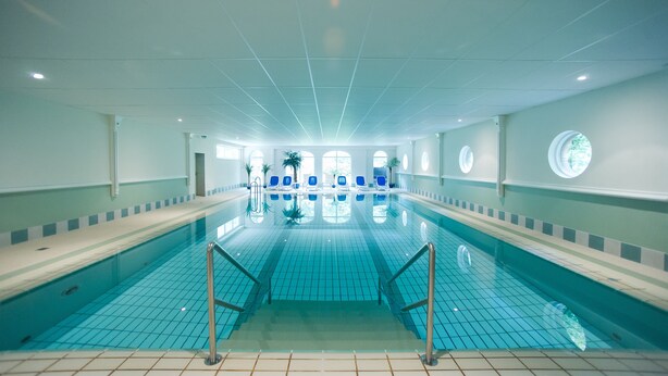 Uma grande piscina interior com água azul cristalina, rodeada por cadeiras azuis e plantas em vasos.