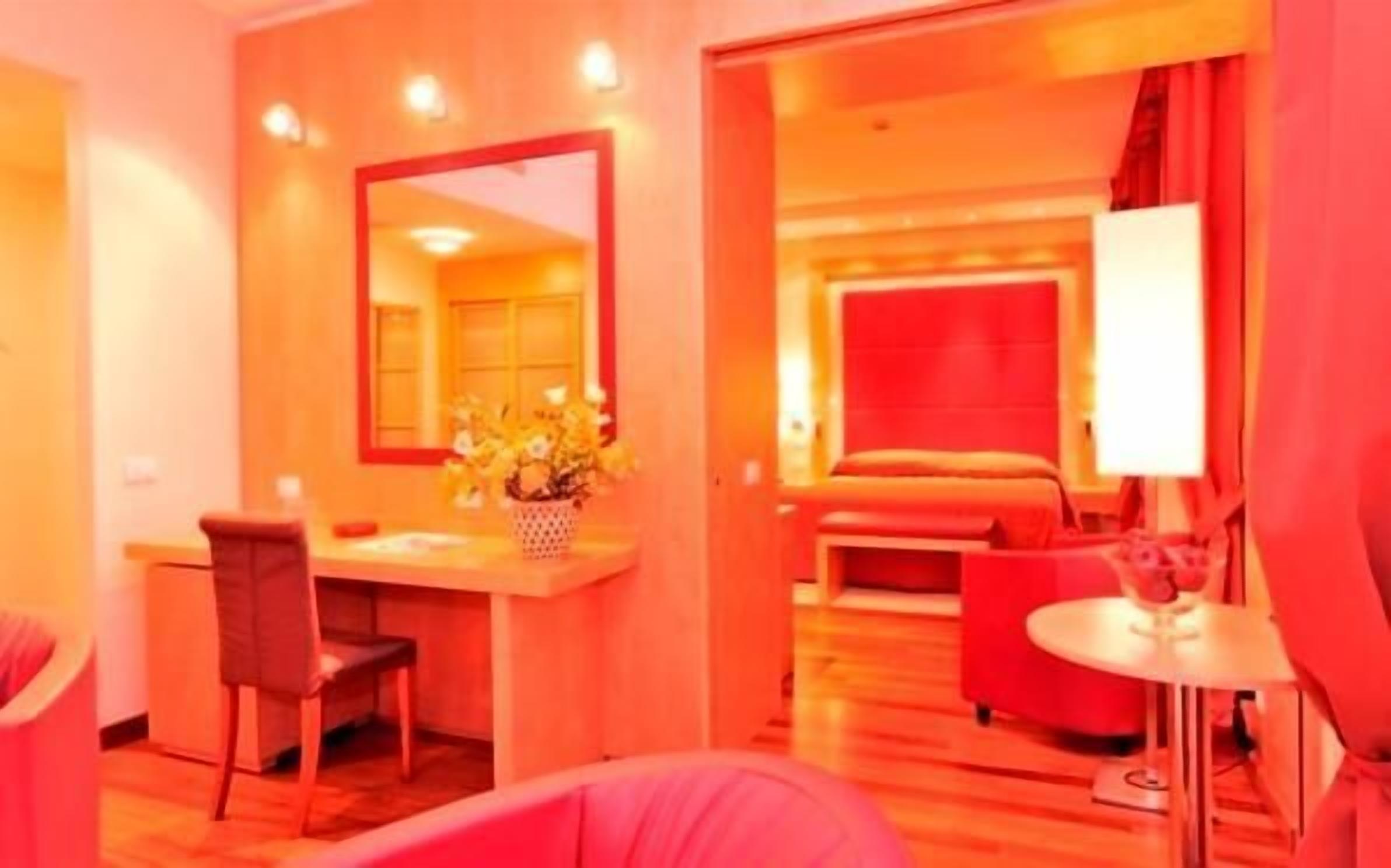Quarto de hotel com secretária de madeira, cadeira e um espelho grande. Há uma cama com roupa de cama vermelha e uma pequena mesa com abajur.