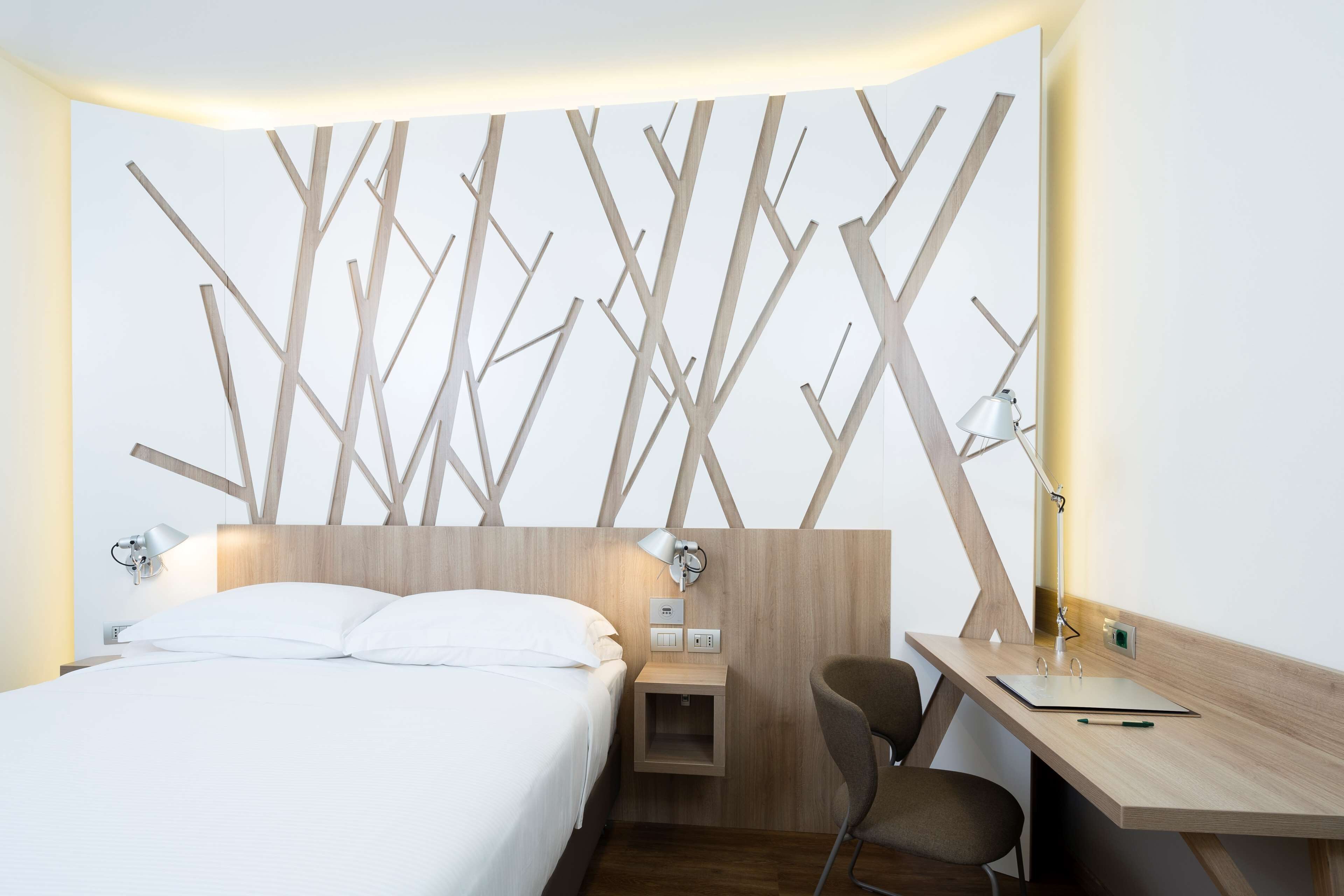 Quarto de hotel moderno com cama, mesa de cabeceira, abajur, escrivaninha, cadeira e uma parede com design geométrico.