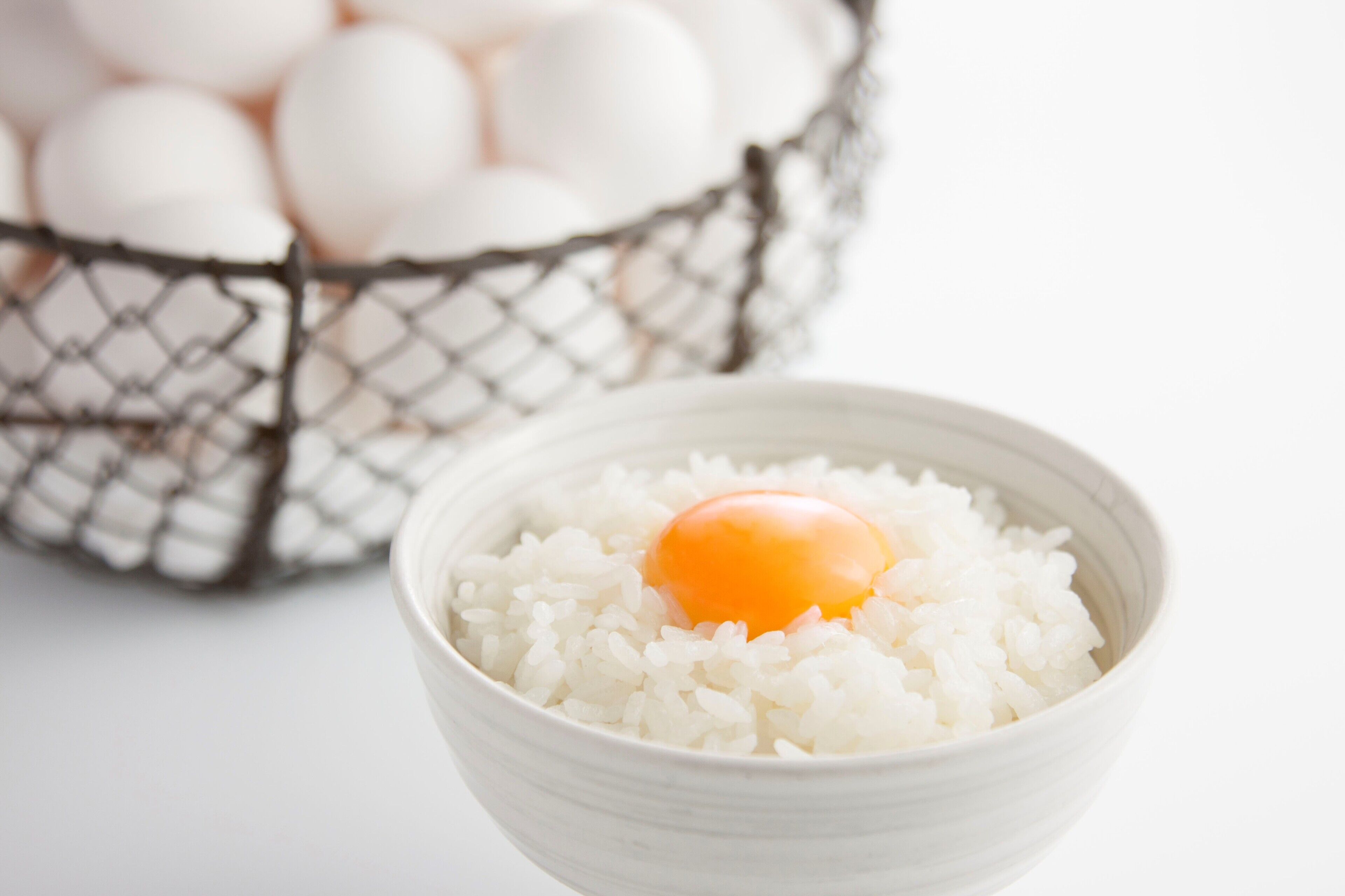Uma tigela de arroz branco com uma gema de ovo por cima, e uma cesta de ovos brancos ao fundo.