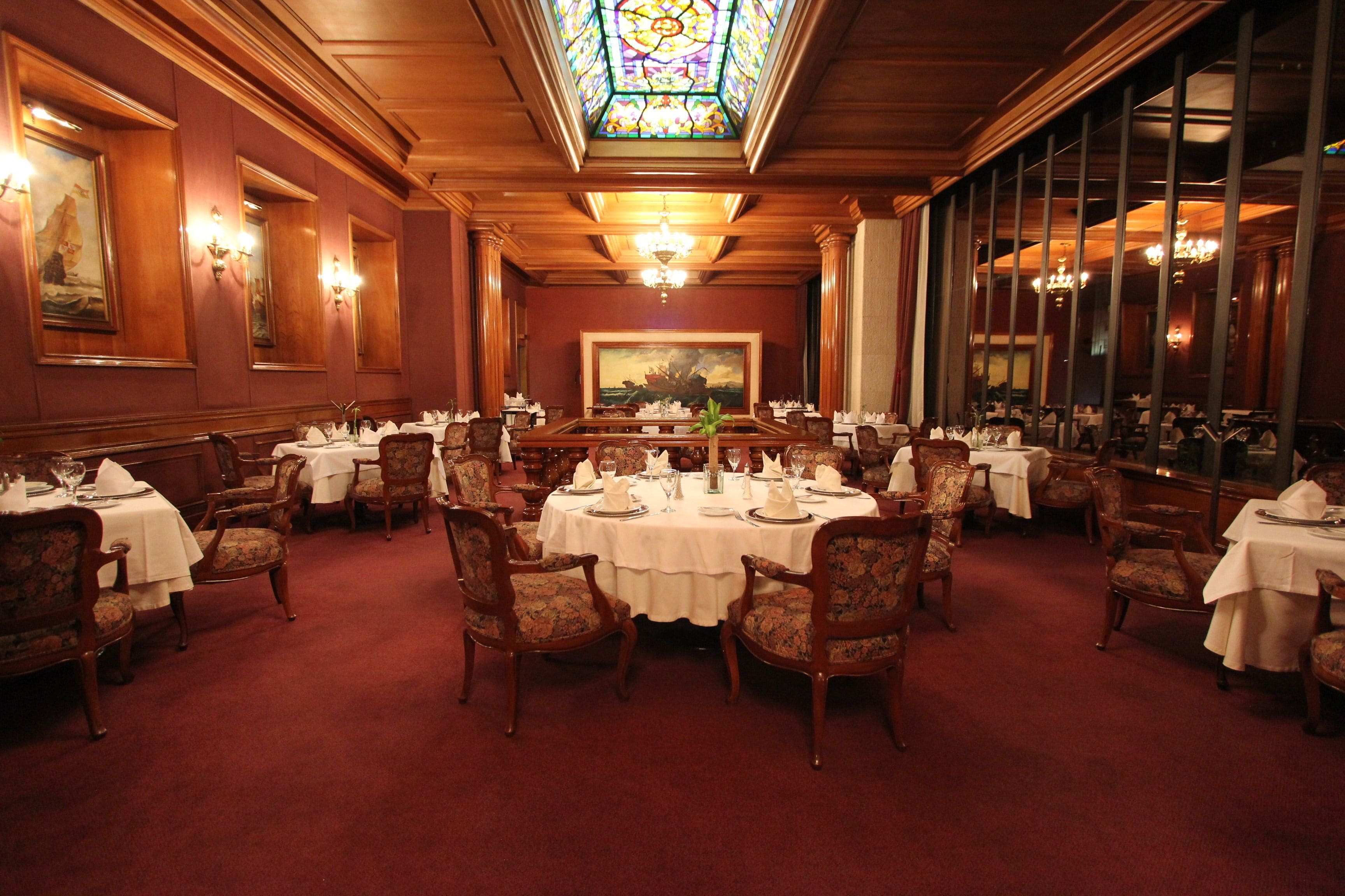 Uma sala de jantar formal com mesas redondas dispostas para vários convidados, painéis de madeira nas paredes e um quadro grande sobre as mesas.