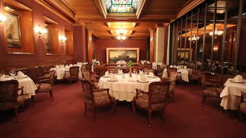 Uma sala de jantar formal com mesas redondas dispostas para vários convidados, painéis de madeira nas paredes e um quadro grande sobre as mesas.