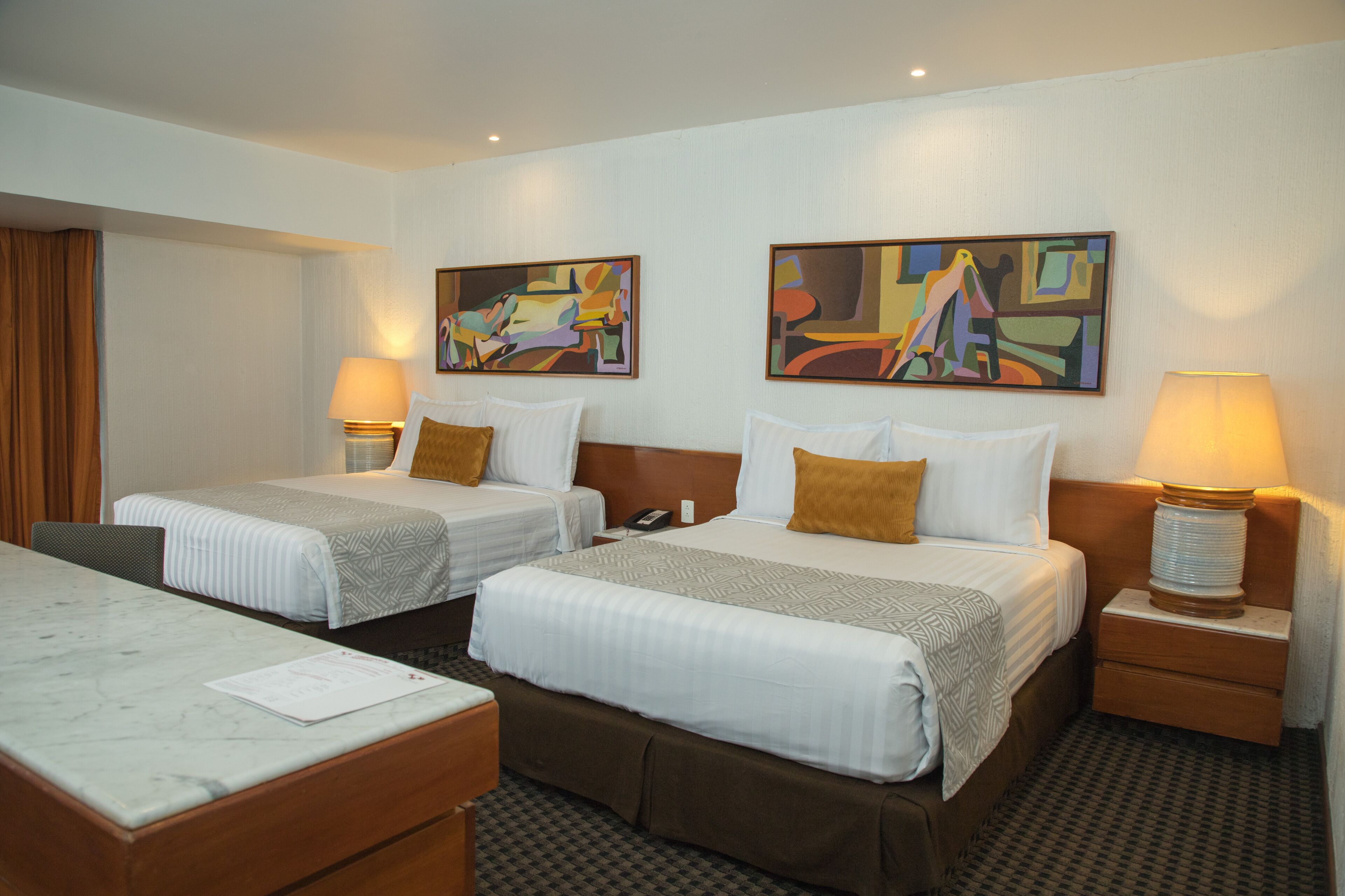 Quarto de hotel com duas camas, uma escrivaninha e uma mesa de cabeceira com abajures. Há quadros abstratos nas paredes.