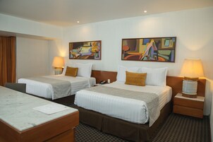 Quarto de hotel com duas camas, uma escrivaninha e uma mesa de cabeceira com abajures. Há quadros abstratos nas paredes.