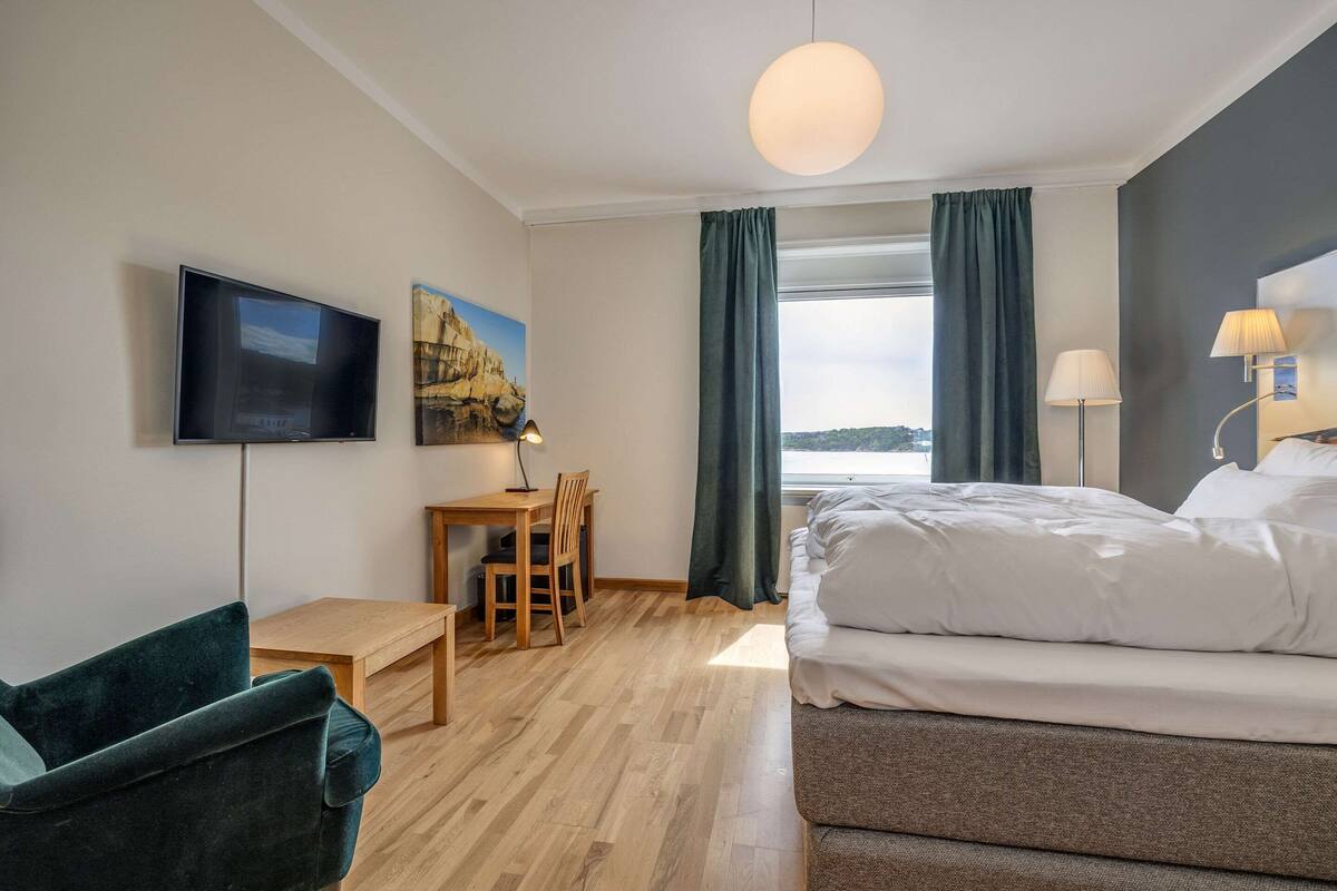 Quarto de hotel com cama, escrivaninha, cadeira, televisão e janela com cortinas.