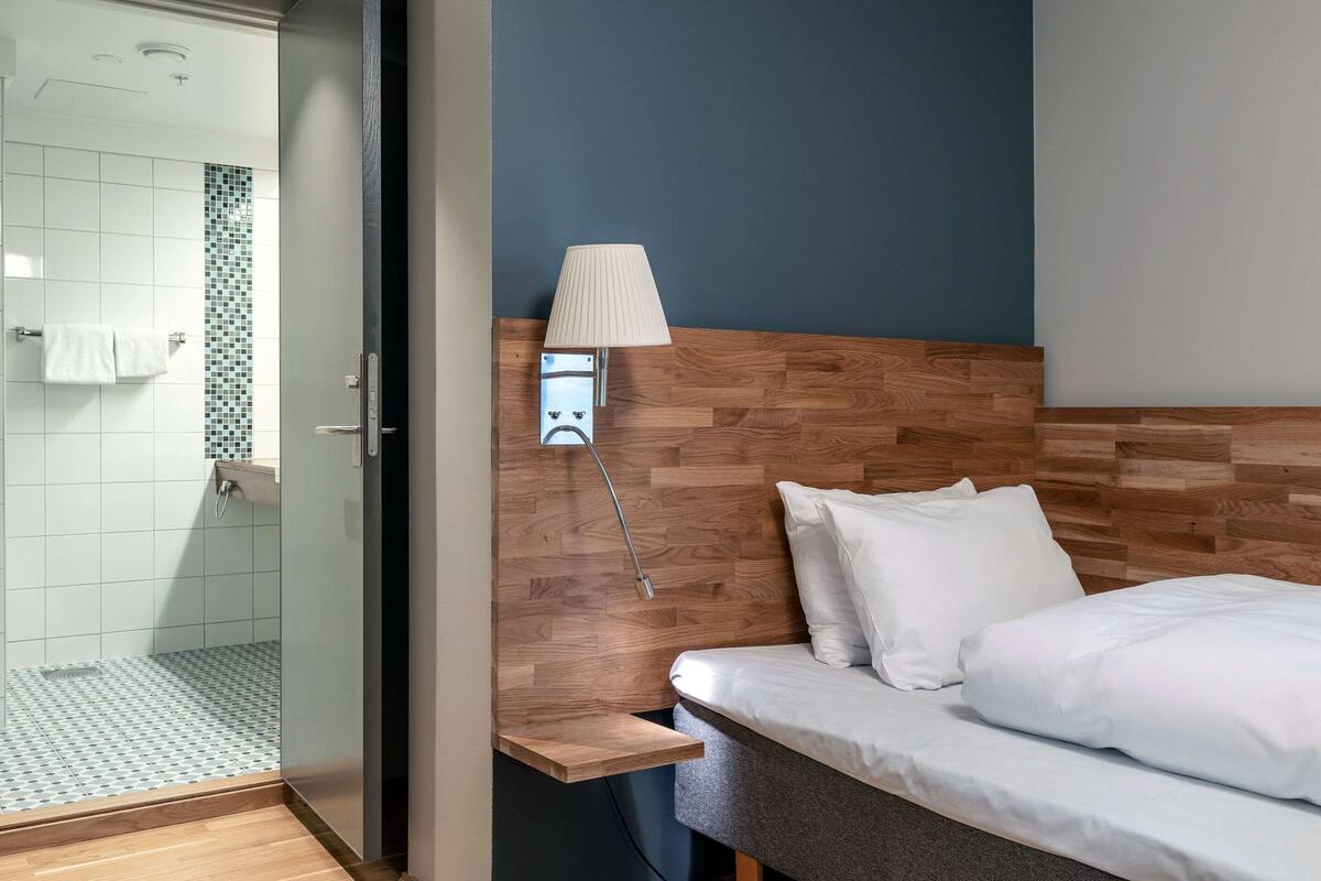 Quarto de hotel com cabeceira de madeira, mesa de cabeceira com luminária, cama com roupa de cama branca e área de chuveiro com piso ladrilhado.