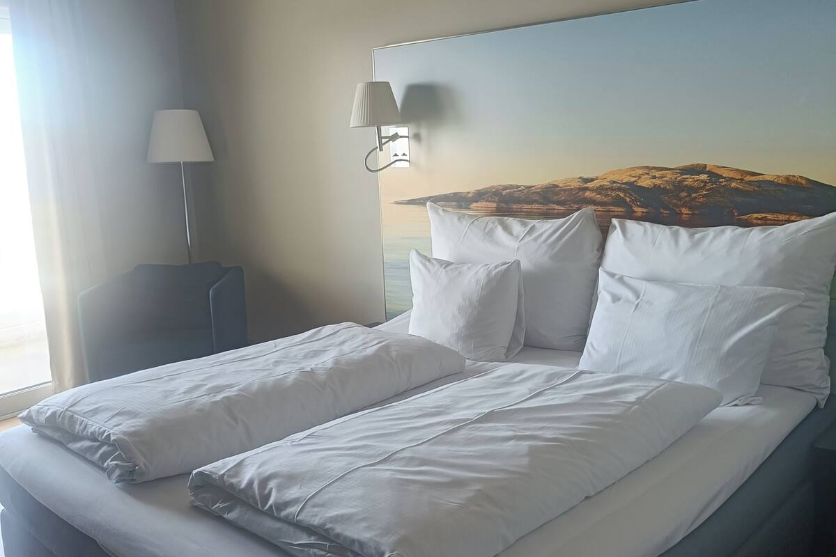 Cama bem arrumada com lençóis brancos em quarto de hotel com uma janela grande, luminária e pintura de paisagem.