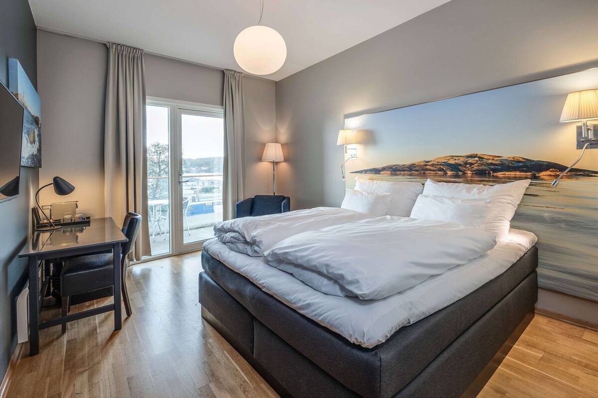 Quarto de hotel com cama, escrivaninha, cadeira e uma grande janela.