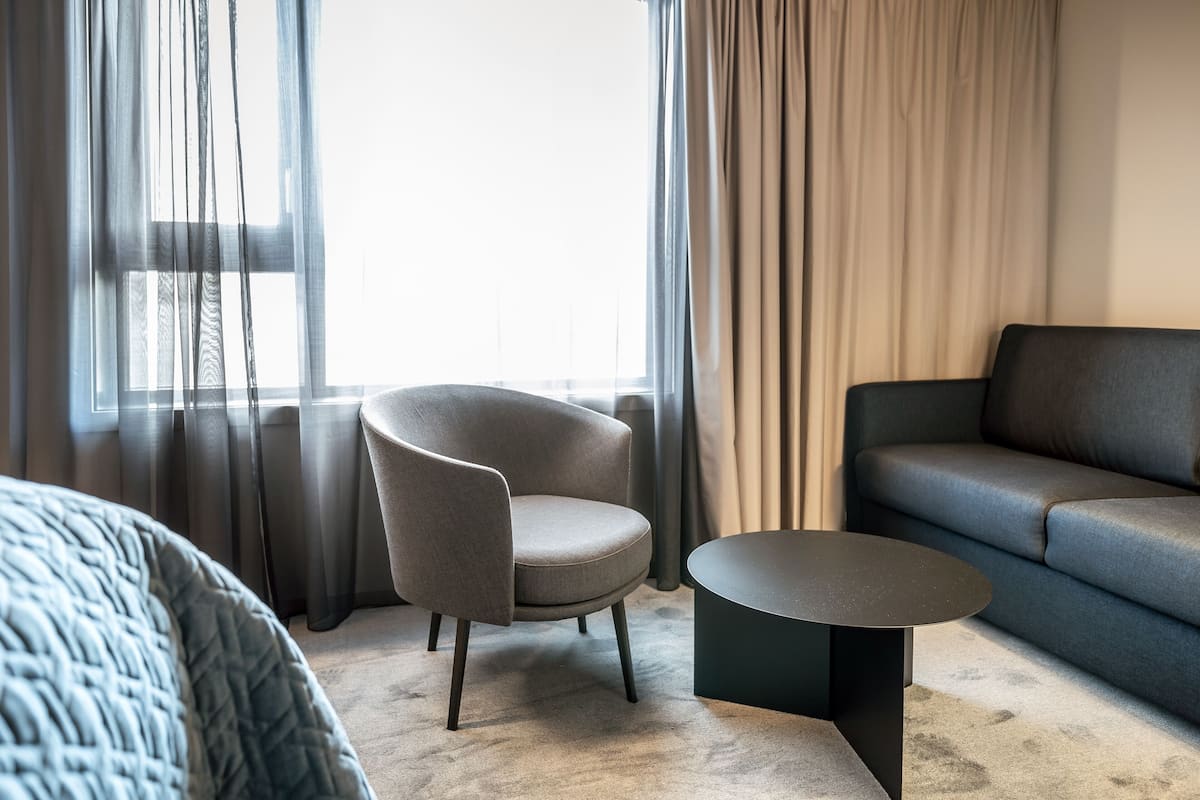 Quarto de hotel moderno com poltrona cinza, sofá escuro e mesa de centro redonda.
