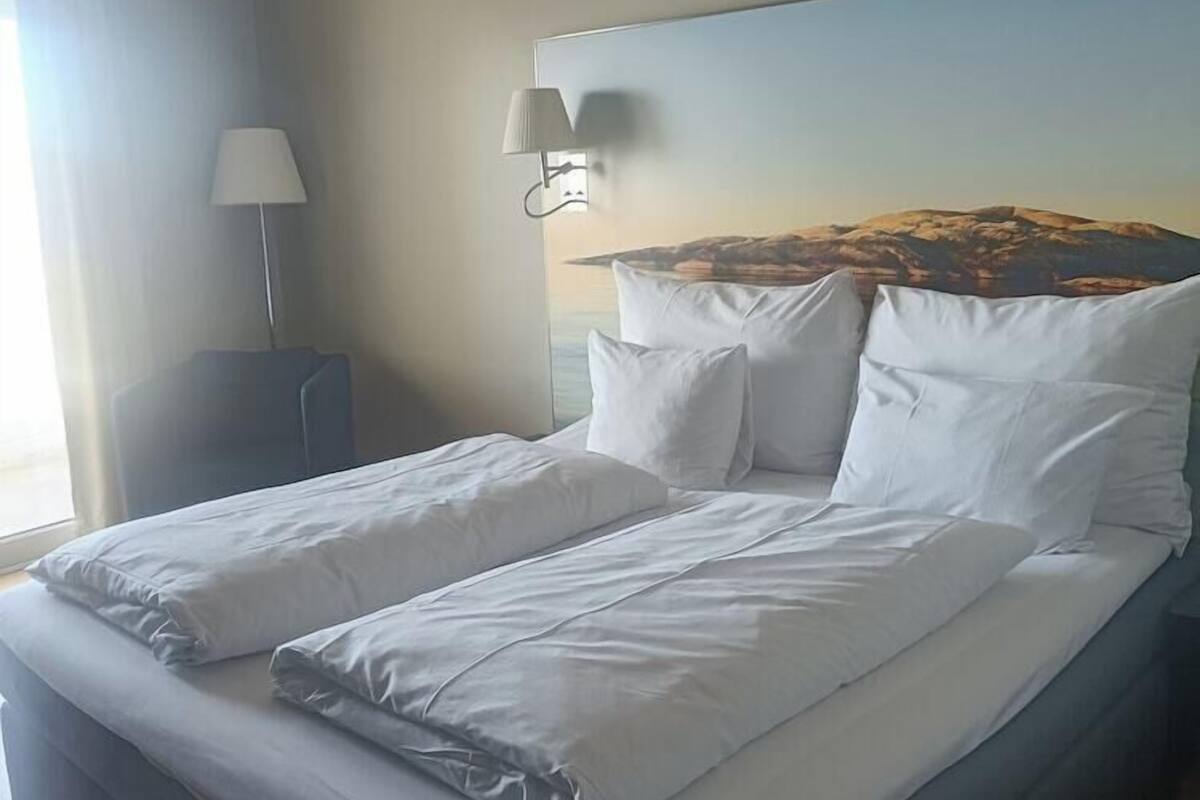 Uma cama bem arrumada com roupa de cama branca, uma luminária e um quadro de uma paisagem na parede.