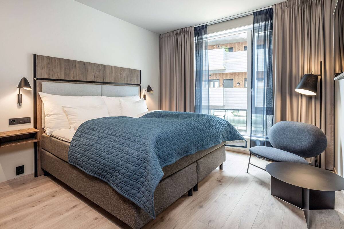 Quarto de hotel moderno com uma cama grande, uma cadeira, uma mesinha e uma janela com cortinas.