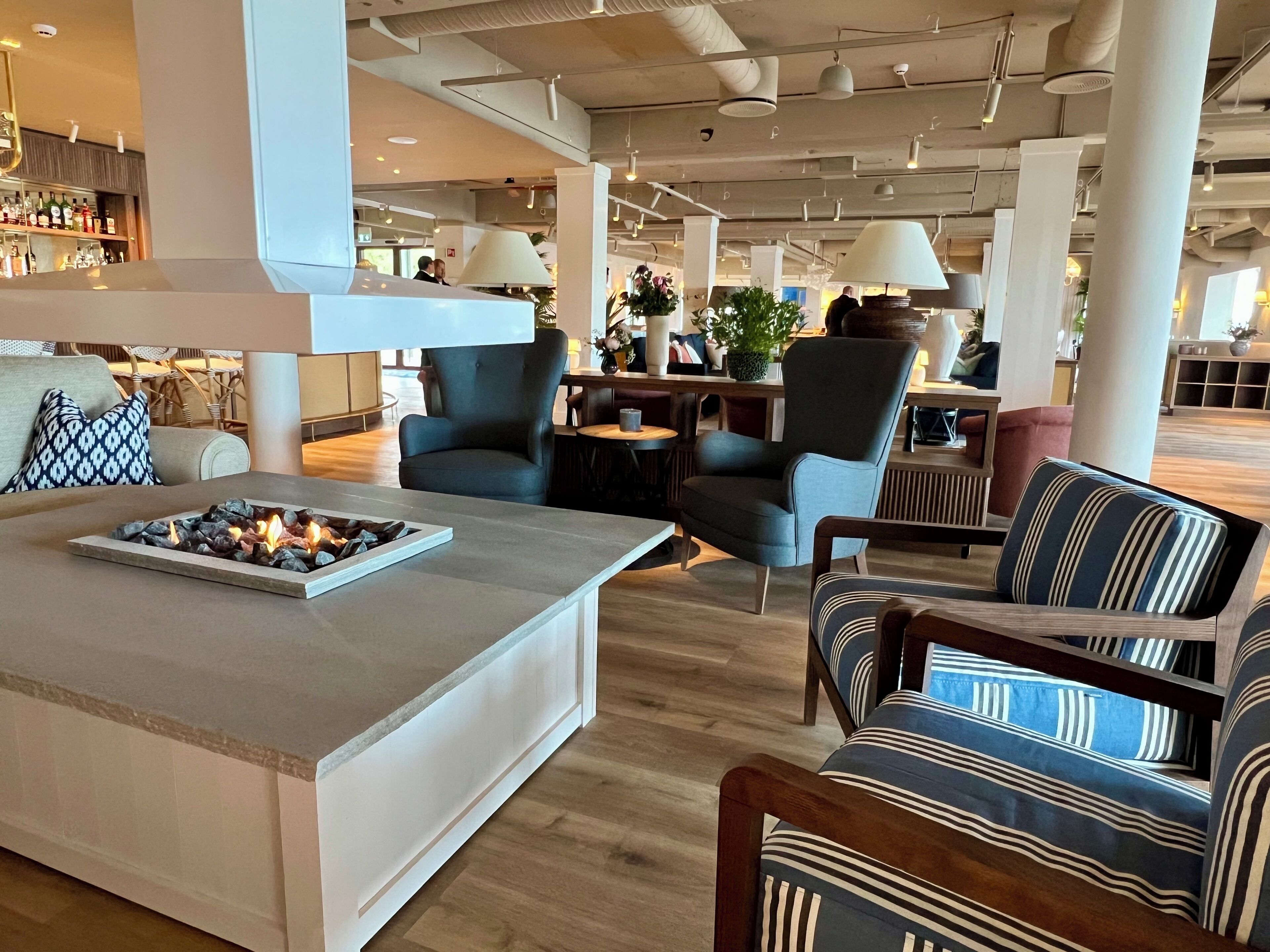 Um hall amplo com área de estar central, apresentando um recurso de fogo, poltronas confortáveis e um bar com diversas garrafas.