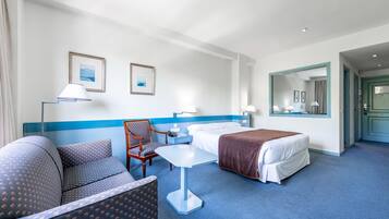 Quarto de hotel com cama, uma cadeira, uma mesinha e duas imagens emolduradas na parede.