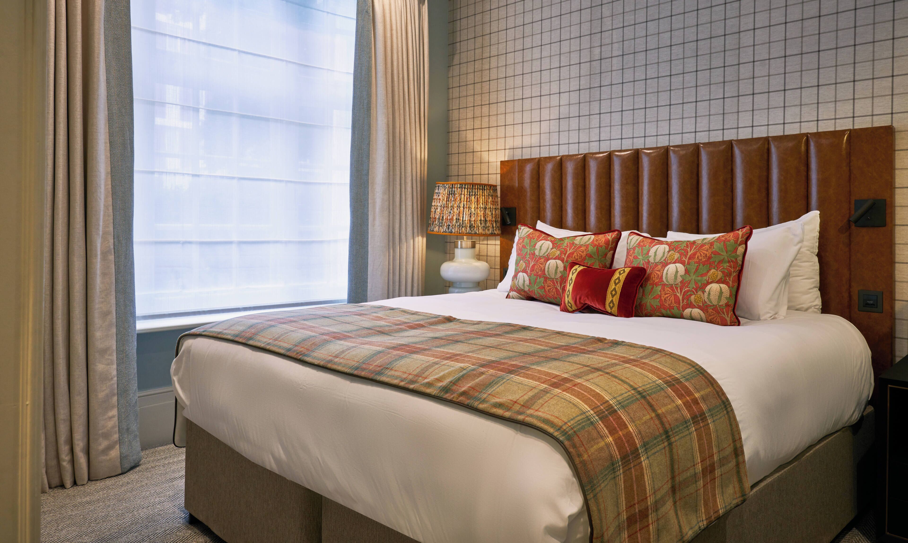 Quarto de hotel com uma cama grande, mesa de cabeceira, luminária e janela com cortinas.