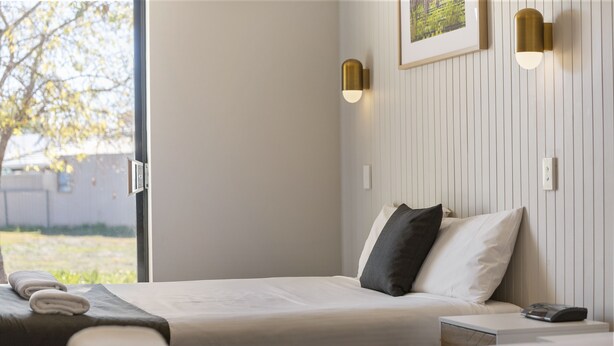 Quarto de hotel com uma cama, mesa de cabeceira, luminária na parede e janela com vista para o verde lá fora.