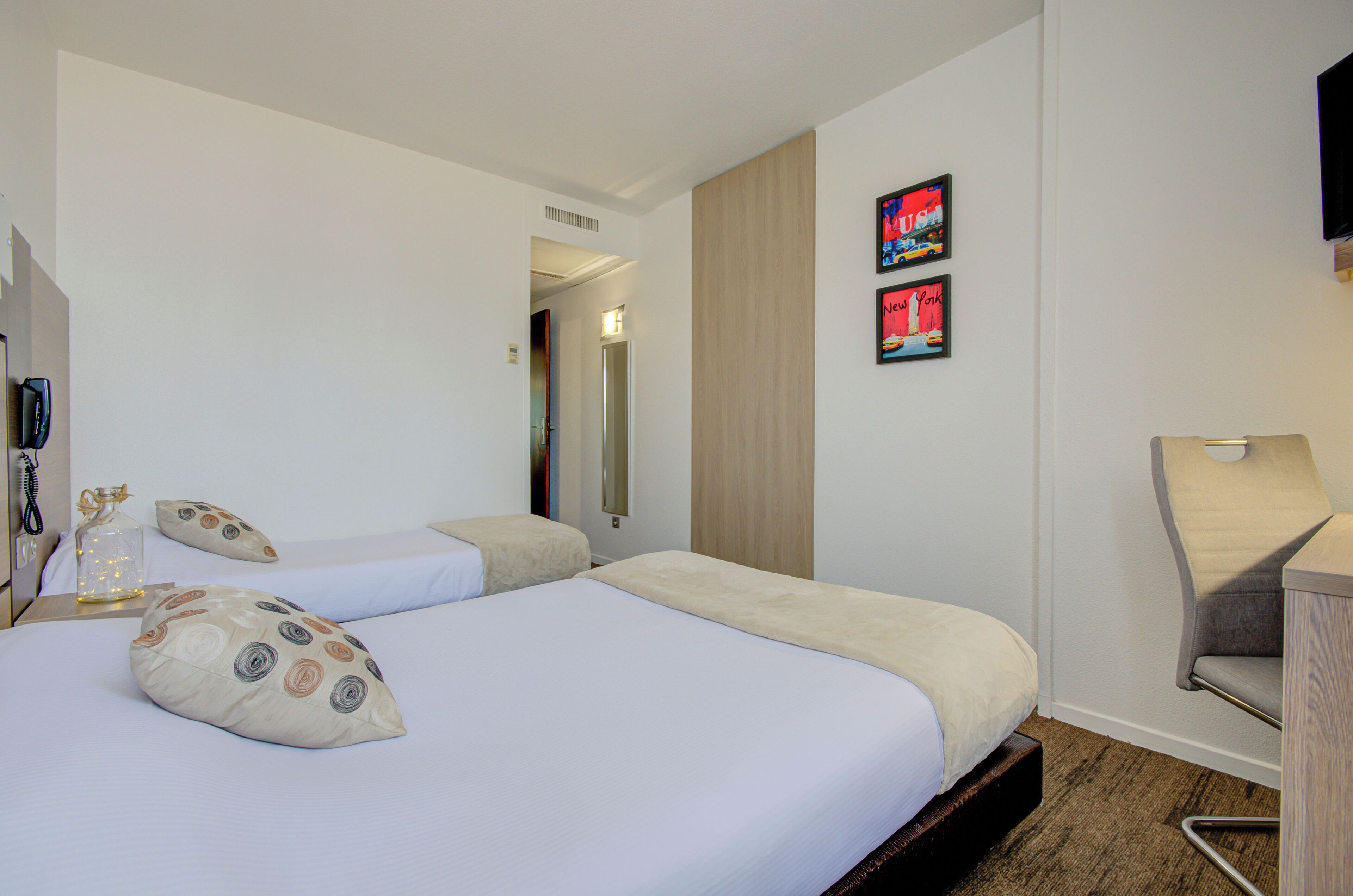 Quarto de hotel com duas camas, uma mesa, uma cadeira e televisão na parede.