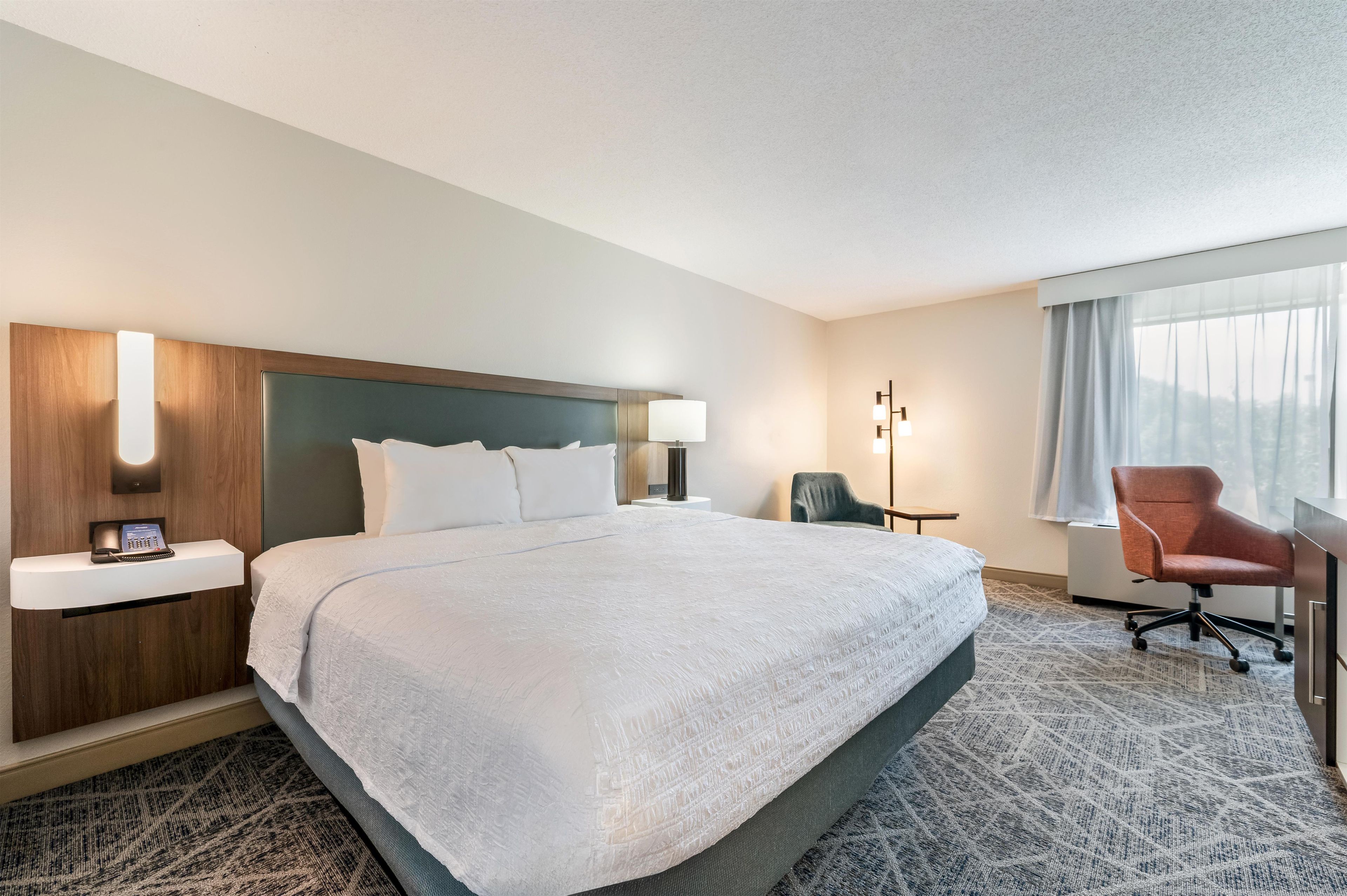 Quarto de hotel com uma cama grande, uma escrivaninha, uma cadeira, um abajur e uma janela com cortinas.