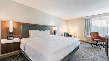 Quarto de hotel com uma cama grande, uma escrivaninha, uma cadeira, um abajur e uma janela com cortinas.