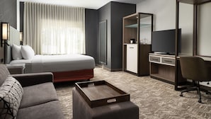 Quarto de hotel moderno com uma cama, um sofá, uma escrivaninha com computador, uma televisão e uma janela grande com cortinas.