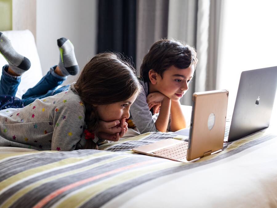 Duas pessoas deitadas em uma cama, com um laptop, usando um smartphone, e uma janela com cortinas ao fundo.