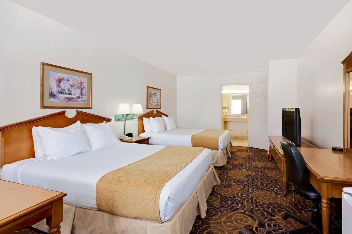 Quarto de hotel com duas camas, uma escrivaninha, uma televisão e um banheiro visível por uma porta aberta.
