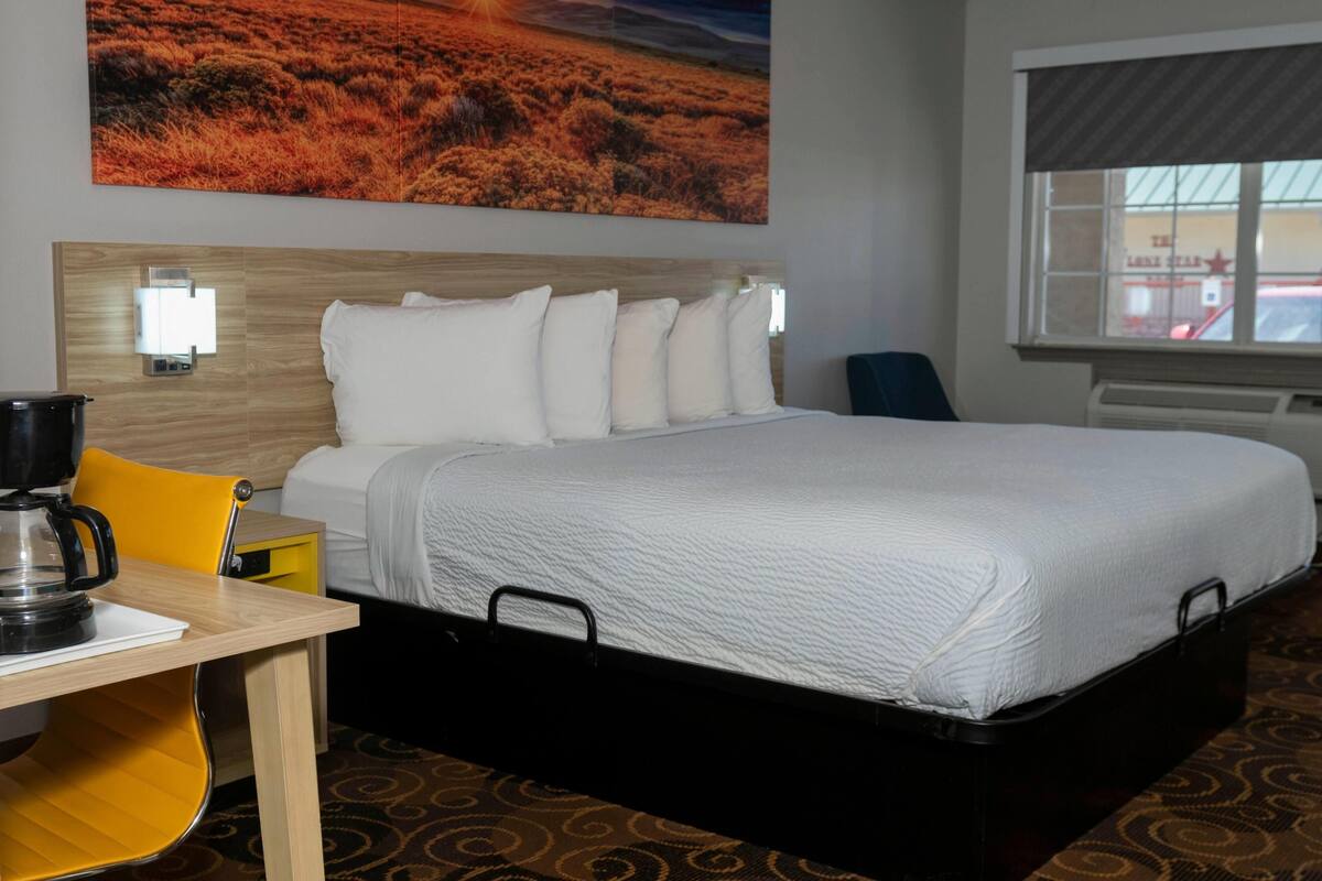 Quarto de hotel com cama, escrivaninha com cafeteira, uma cadeira, quadro na parede e janela com vista para um edifício.