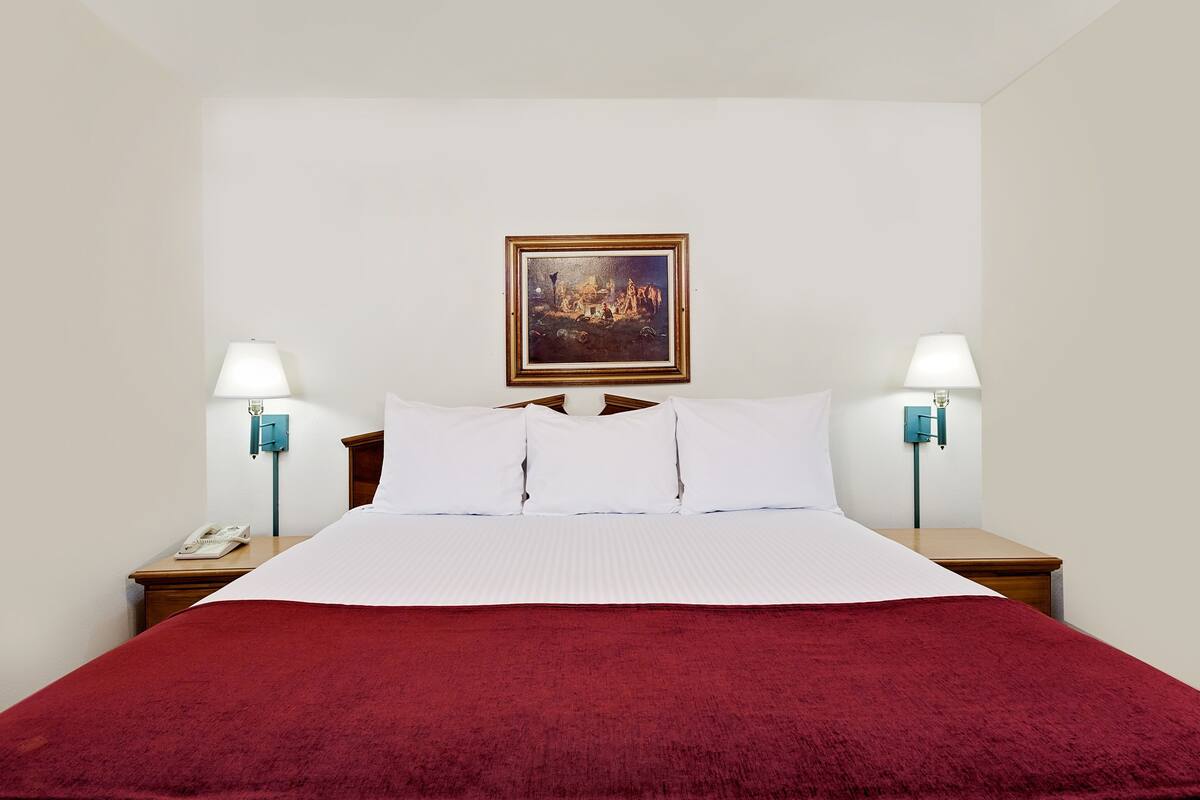 Quarto de hotel com uma cama grande, roupa de cama branca, um cobertor vermelho, duas luminárias de cabeceira, mesa de cabeceira com telefone e um quadro na parede.