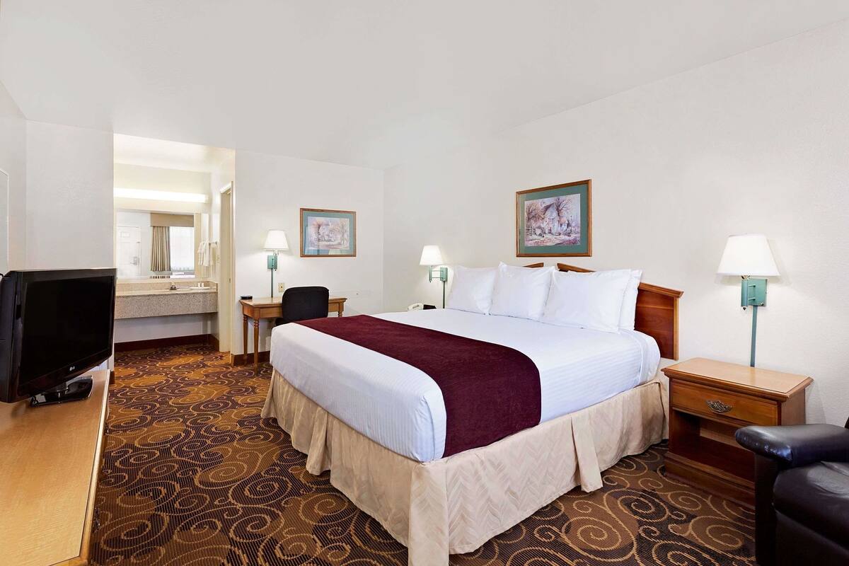 Quarto de hotel com cama, escrivaninha com televisão, cadeira, mesa de cabeceira e abajur.