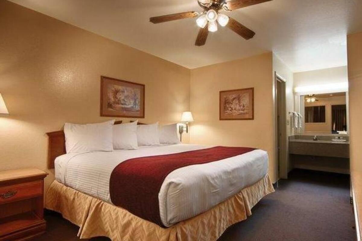 Quarto de hotel com uma cama, um ventilador de teto, duas luminárias fixadas na parede, um espelho e uma porta que dá para um banheiro.