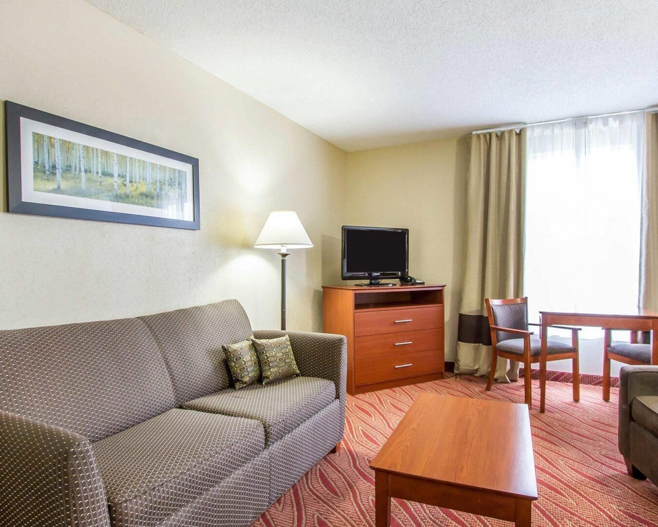 Quarto de hotel com sofá, mesa de madeira, televisão em móvel de madeira, luminária, cadeira e janela com cortinas.
