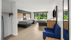 Quarto de hotel moderno com três camas, uma poltrona azul, uma escrivaninha de madeira e uma janela grande com vista para o verde.