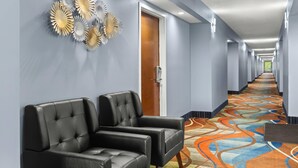 Um corredor de hotel com duas poltronas de couro preto, um objeto decorativo na parede e um tapete estampado.