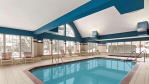 Piscina interna com água azul cristalina, rodeada por cadeiras e mesas brancas, grandes janelas e teto alto com vigas azuis.