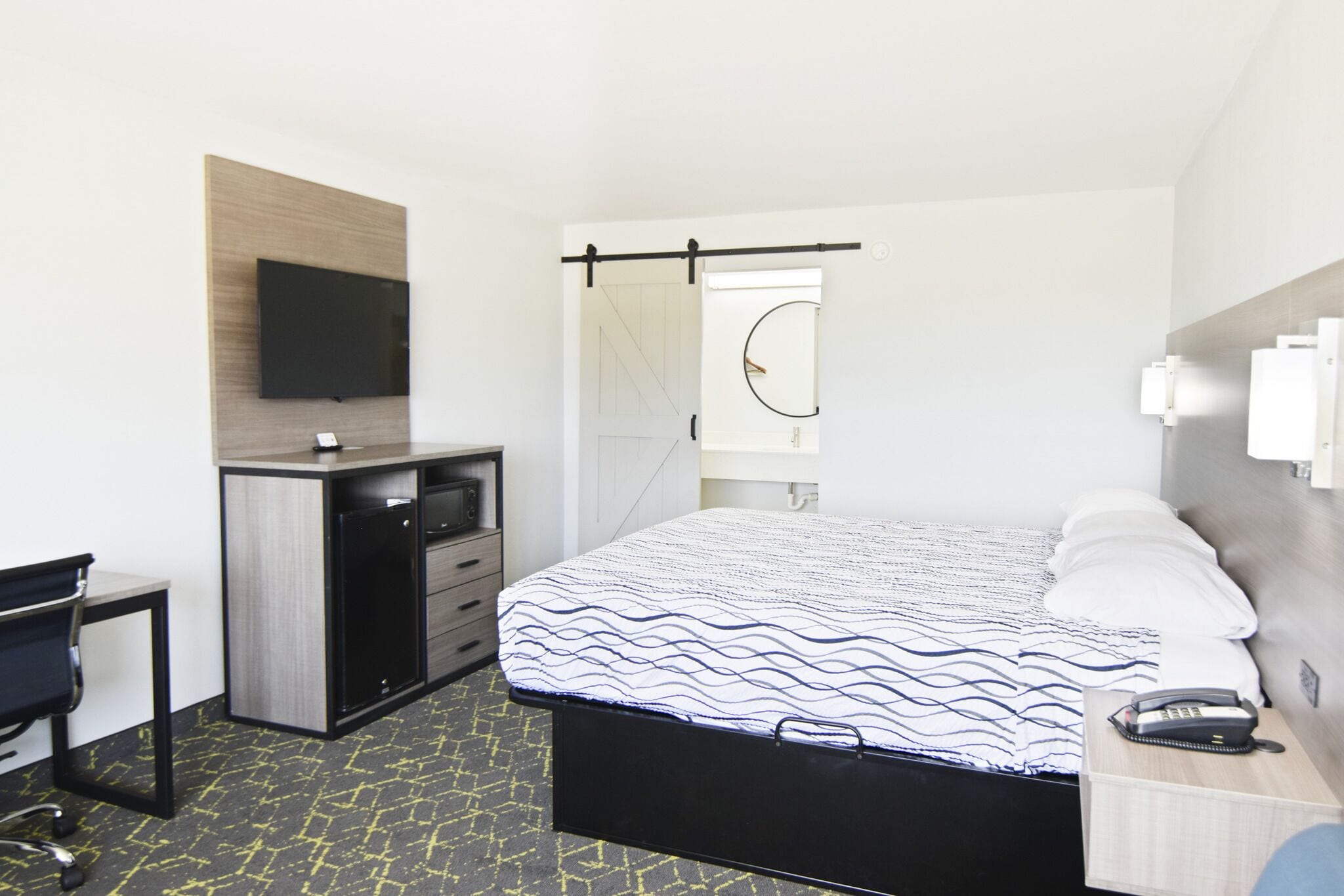Quarto de hotel com cama, escrivaninha, televisão na parede, banheiro com espelho e telefone na mesa de cabeceira.